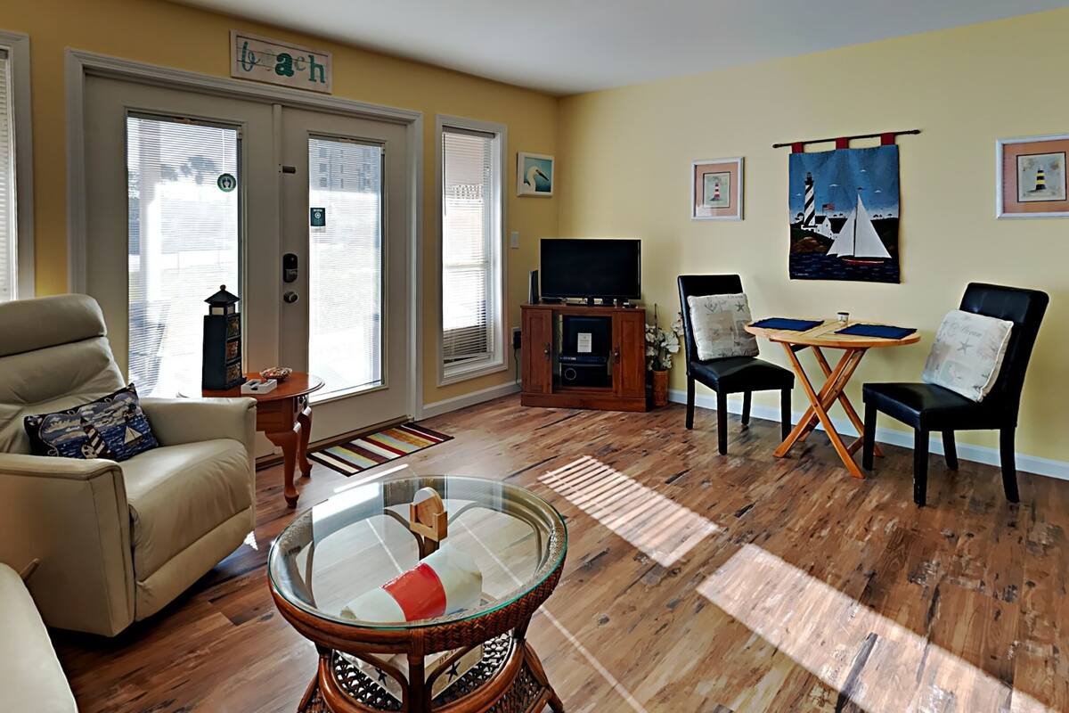 Uma sala de estar com um sofá bege, uma mesa de centro de vidro, um rack para TV e um quadro com barcos à vela.