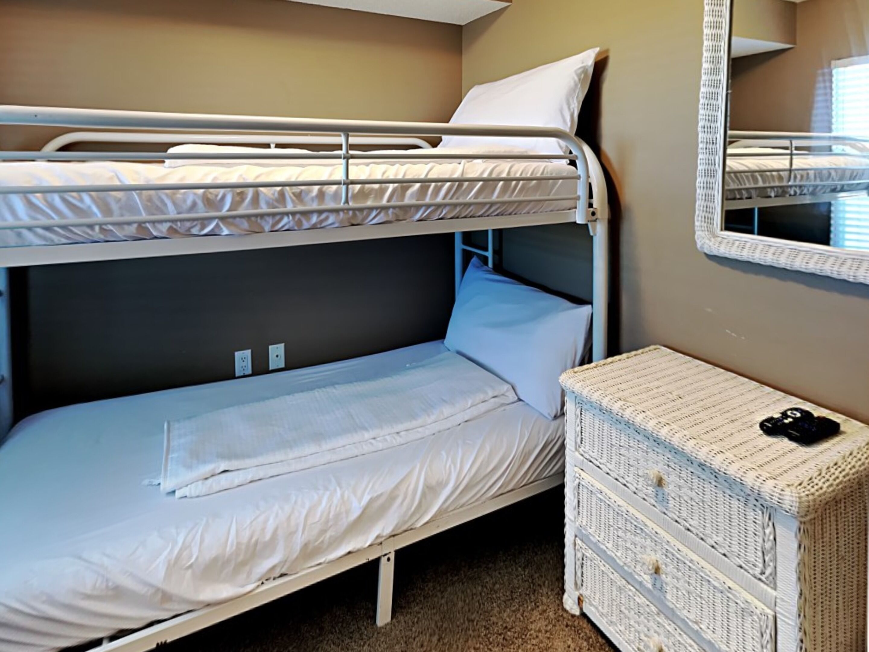 Quarto com beliche, colchão branco, cabeceira branca, mesa de cabeceira pequena branca com luminária e espelho na parede.