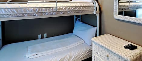 Quarto com beliche, colchão branco, cabeceira branca, mesa de cabeceira pequena branca com luminária e espelho na parede.