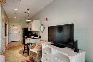 Uma sala de estar com televisão, mobiliário branco e uma cozinha ao fundo.