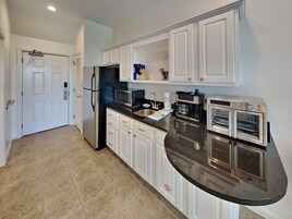 Cozinha compacta com armários brancos, bancada preta, geladeira, micro-ondas, cafeteira e forno elétrico.