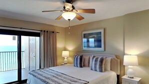 Quarto de hotel com uma cama, duas mesas de cabeceira com luminárias, um ventilador de teto, uma janela grande com cortinas e um quadro na parede.
