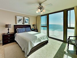 Quarto com uma cama grande, um ventilador de teto e vista para o oceano através de amplas janelas.