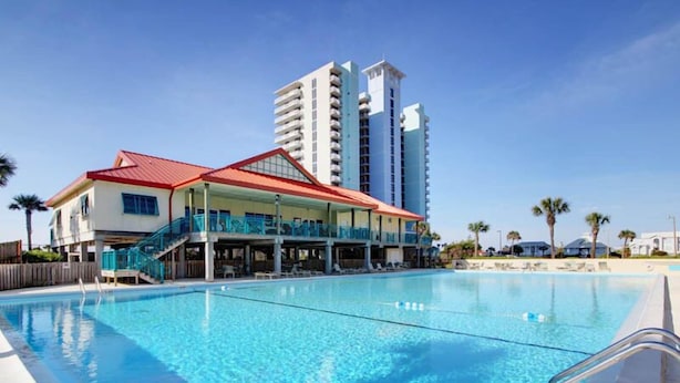 Uma piscina com superfície de água azul cristalina, um edifício com telhado vermelho e varandas, e palmeiras ao fundo.
