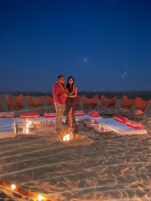 Um casal em pé na praia, com cadeiras e mesas dispostas para um evento ao ar livre.