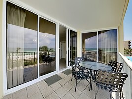 Uma varanda com porta de vidro que dá para um terraço com mesa e cadeiras, com vista para uma praia.