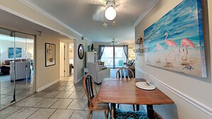 Uma sala de jantar com mesa de madeira, cadeiras e uma cesta de palha. Há um ventilador de teto, um quadro de flamingos e vista para outro quarto com uma cama e uma janela.