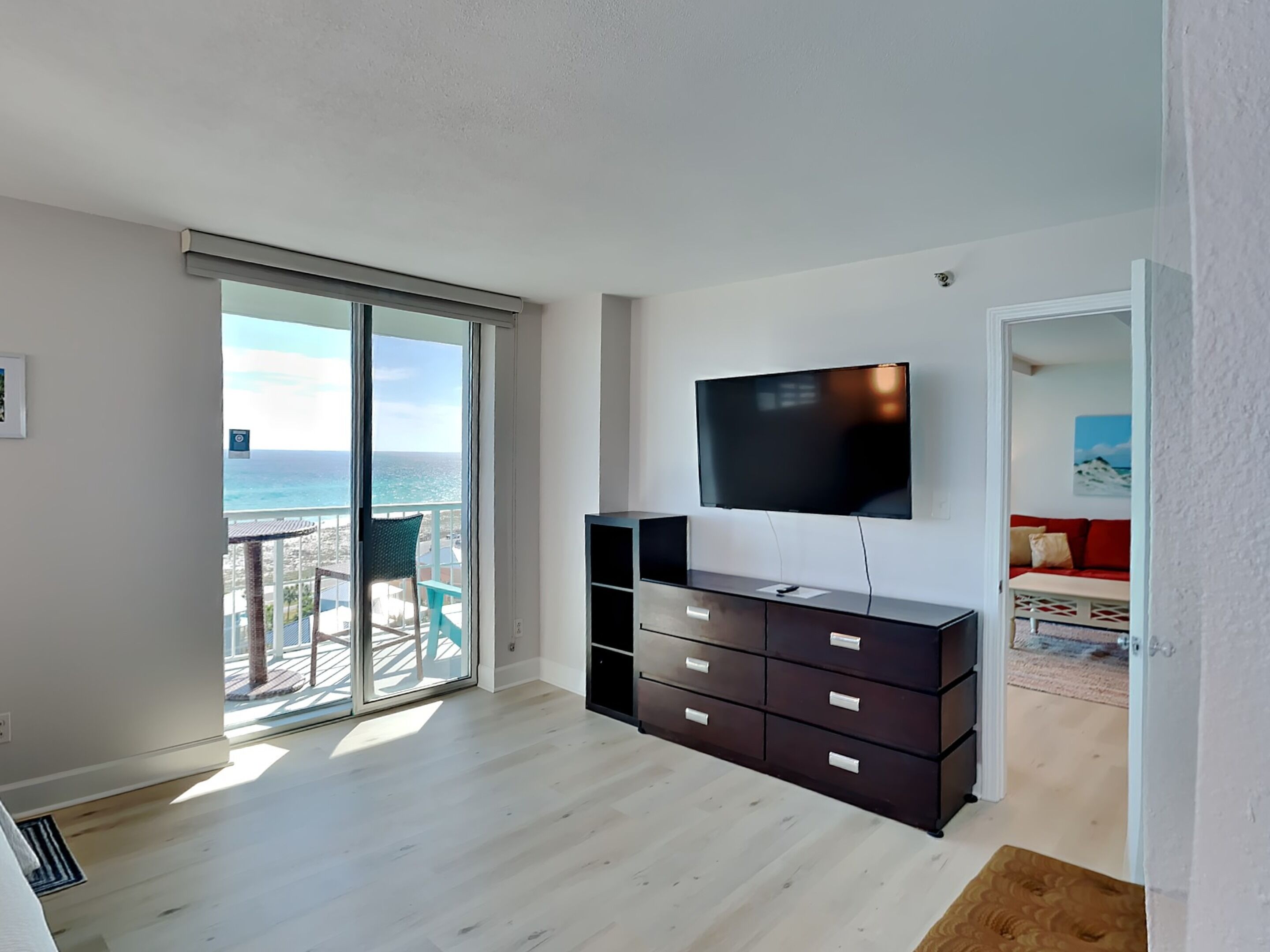 Uma sala de estar moderna com uma televisão de tela plana grande, uma porta de vidro deslizante que dá para uma varanda com vista para o oceano, e um sofá vermelho no canto.