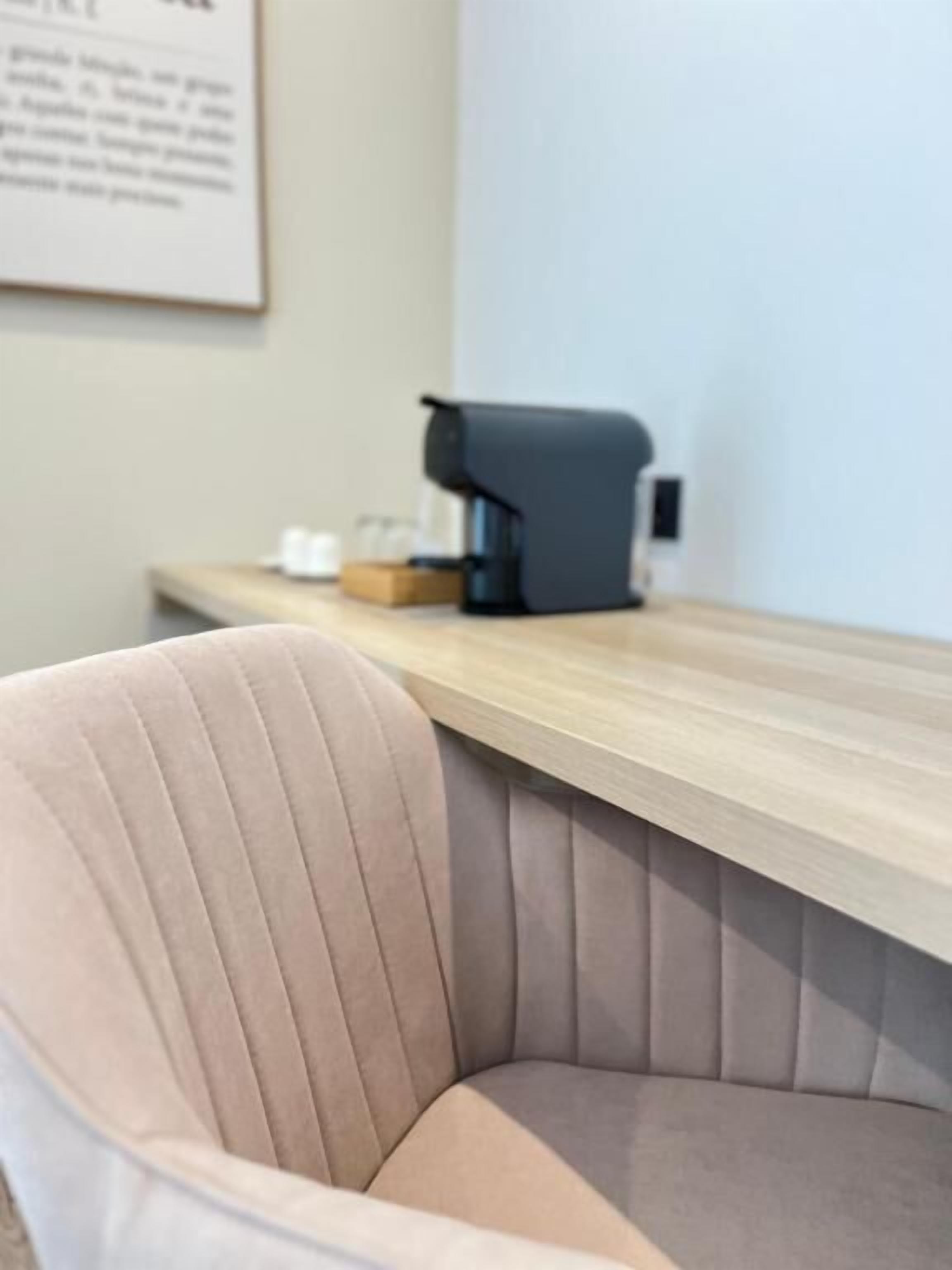 Quarto moderno e minimalista com uma poltrona bege estofada, uma mesa de cabeceira de madeira e uma máquina de café preta.
