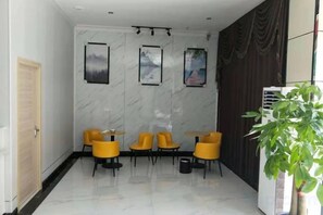 Um saguão de hotel com parede de mármore, quadros emoldurados, cadeiras amarelas e um vaso de planta.