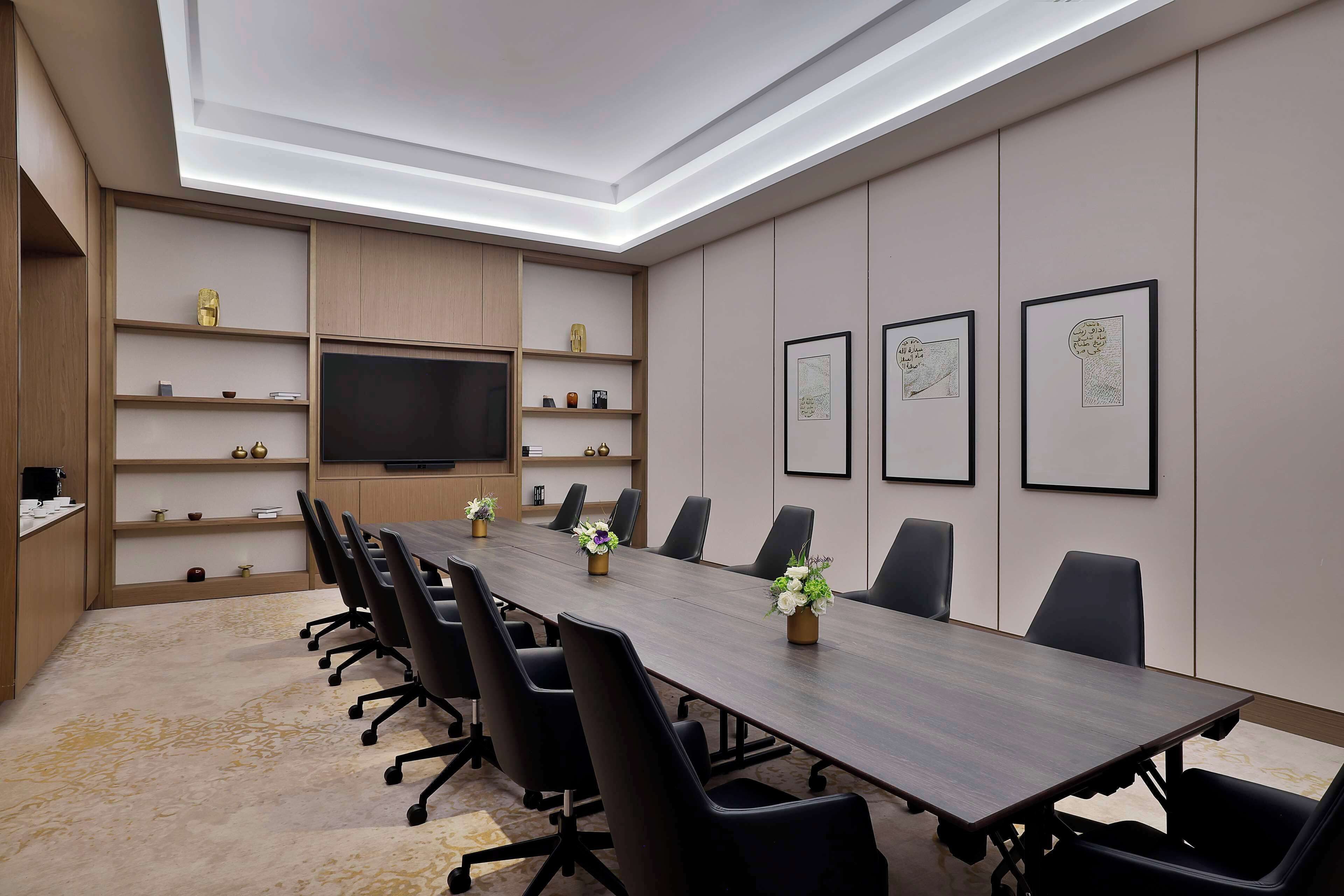 Uma sala de conferências com uma mesa comprida, cadeiras pretas, uma TV, prateleiras com objetos decorativos e quadros nas paredes.