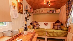 Um quarto aconchegante com teto de madeira, um sofá com almofadas xadrez, uma mesa pequena com taças de vinho e uma garrafa, e uma prateleira na parede com livros e um globo.