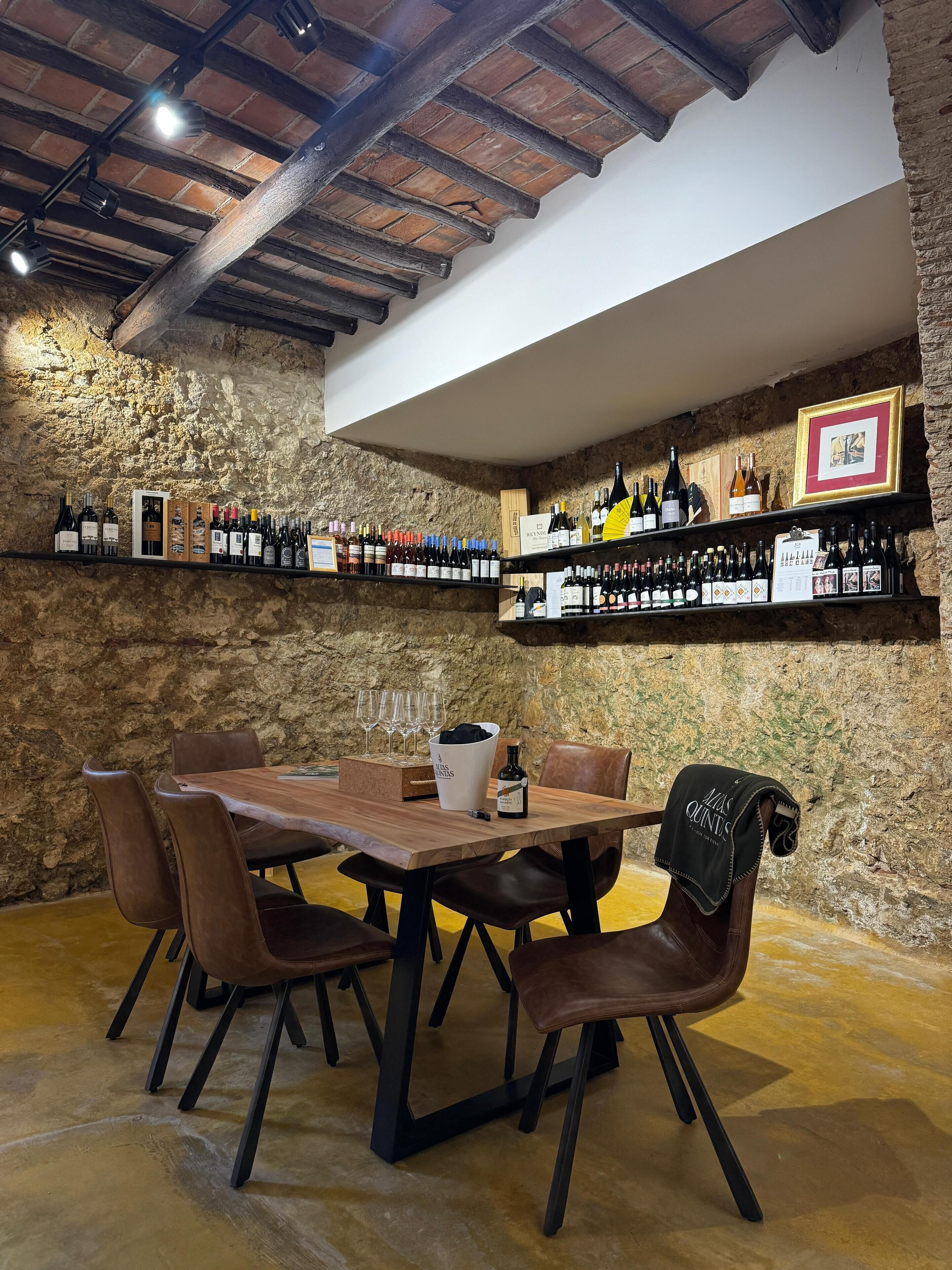 Uma adega rústica com mesas e cadeiras de madeira, prateleiras cheias de garrafas de vinho e um fundo de parede de pedra.