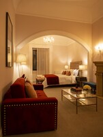 Quarto de hotel com sofá vermelho, mesa de centro de vidro, cama com roupa de cama branca e um candelabro.