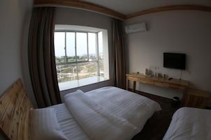 Quarto de hotel com duas camas, uma janela grande com cortinas, cabeceira de madeira, uma pequena mesa com televisão de tela plana e um aparelho de ar condicionado.