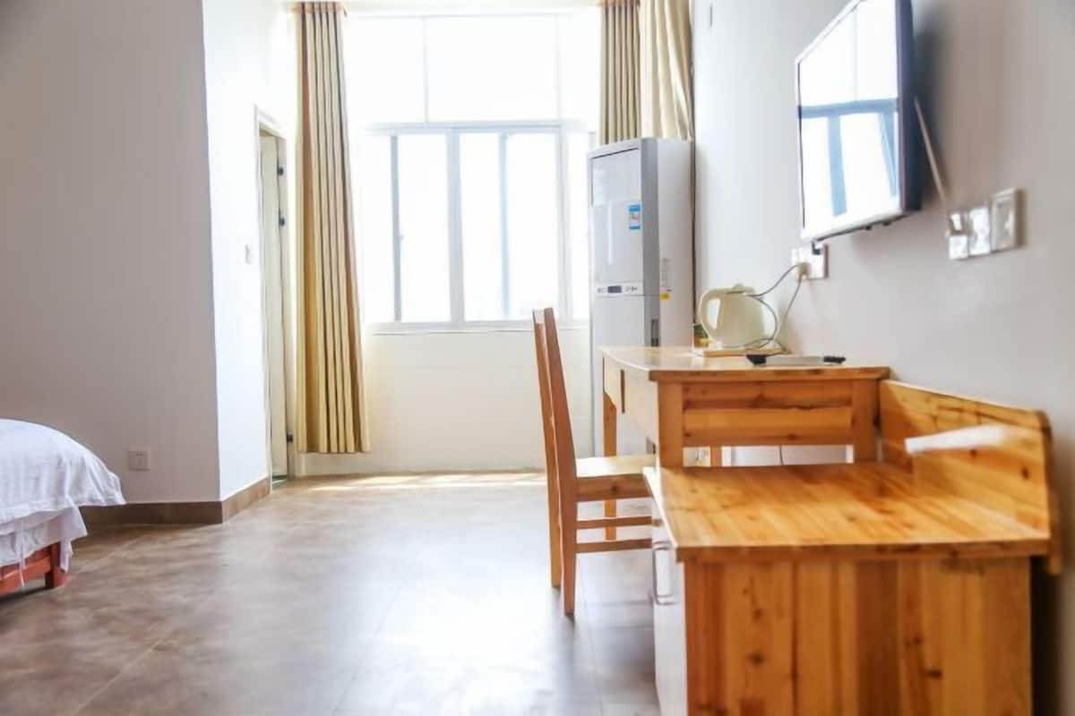 Um quarto com uma cama, uma mesa de madeira, uma cadeira, uma televisão e uma janela com cortinas.