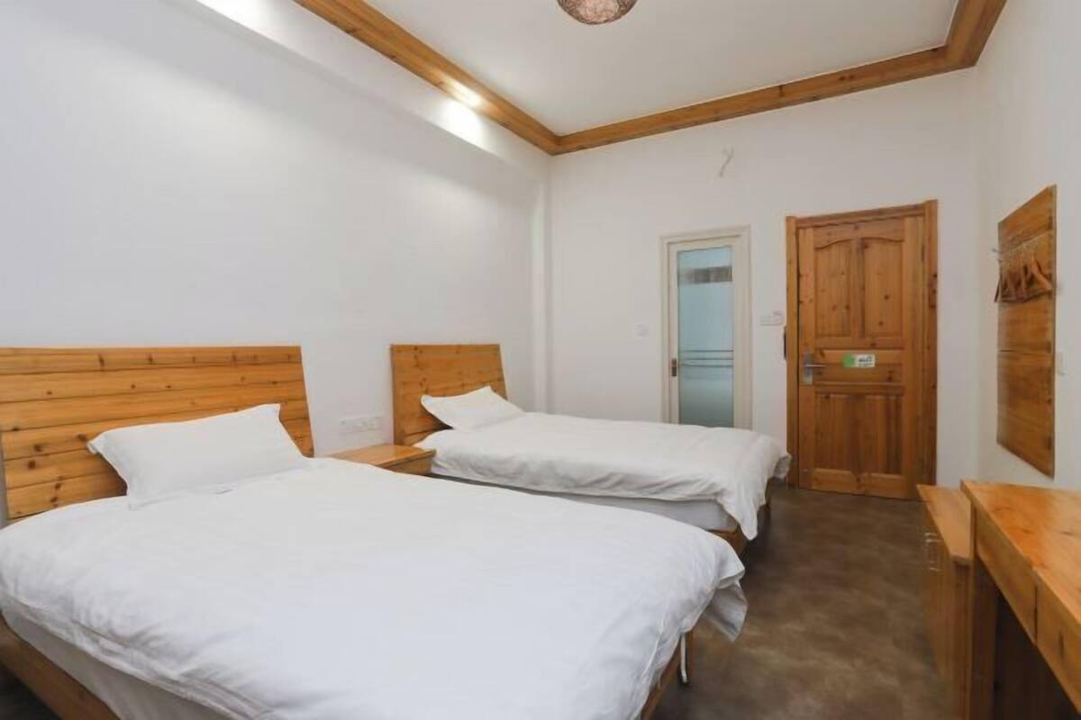 Quarto com duas camas de solteiro, cabeceiras de madeira, porta de madeira e uma pequena mesa de madeira.