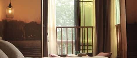 Quarto com cama, luminária pendente, janela com cortinas e vista para o verde lá fora.