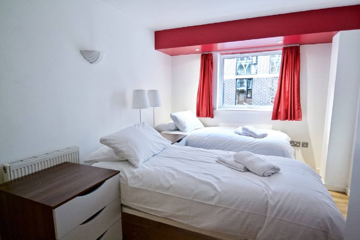 Quarto com duas camas, penteadeira de madeira, radiador e janela com cortinas vermelhas.