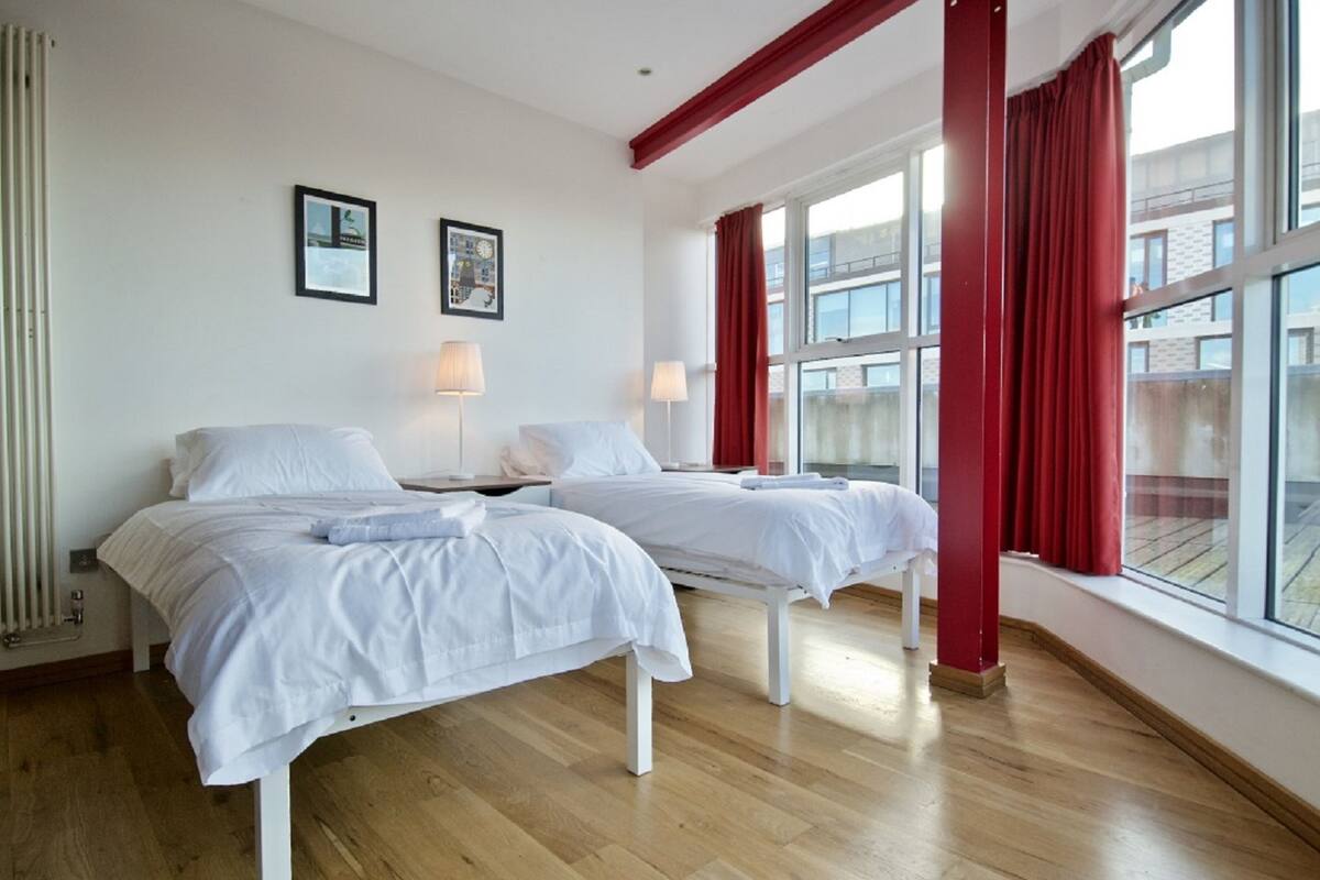 Quarto de hotel com duas camas, uma janela grande com cortinas vermelhas e um radiador na parede.