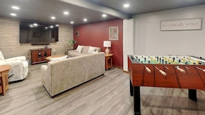 Um quarto aconchegante com sofá, mesa de pebolim, TV e um letreiro na parede.