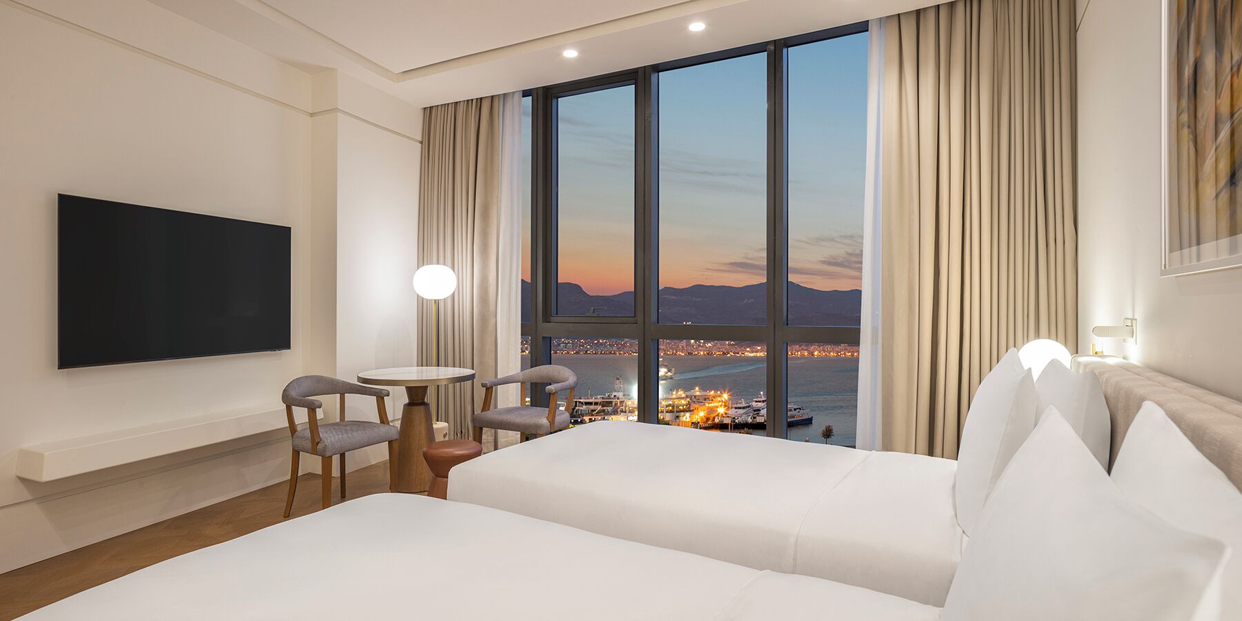 Quarto de hotel com duas camas, televisão, mesa com cadeiras, luminária e vista para a cidade ao pôr do sol.