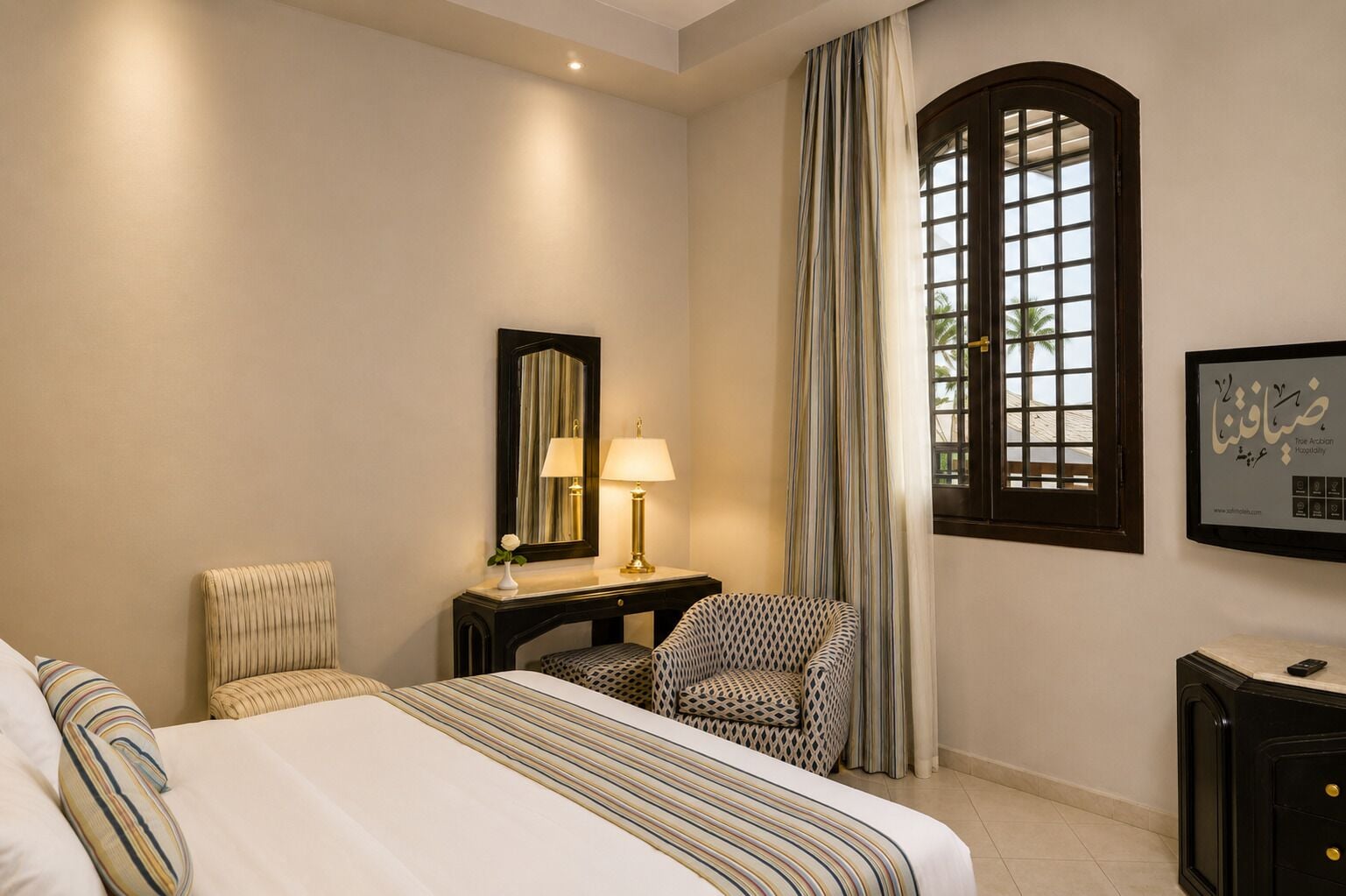 Quarto de hotel com cama, escrivaninha com abajur, uma cadeira, janela com cortinas e um quadro na parede.