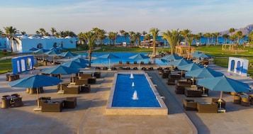 Uma área de piscina em resort com várias espreguiçadeiras e guarda-sóis, uma piscina central com uma fonte e palmeiras ao fundo.