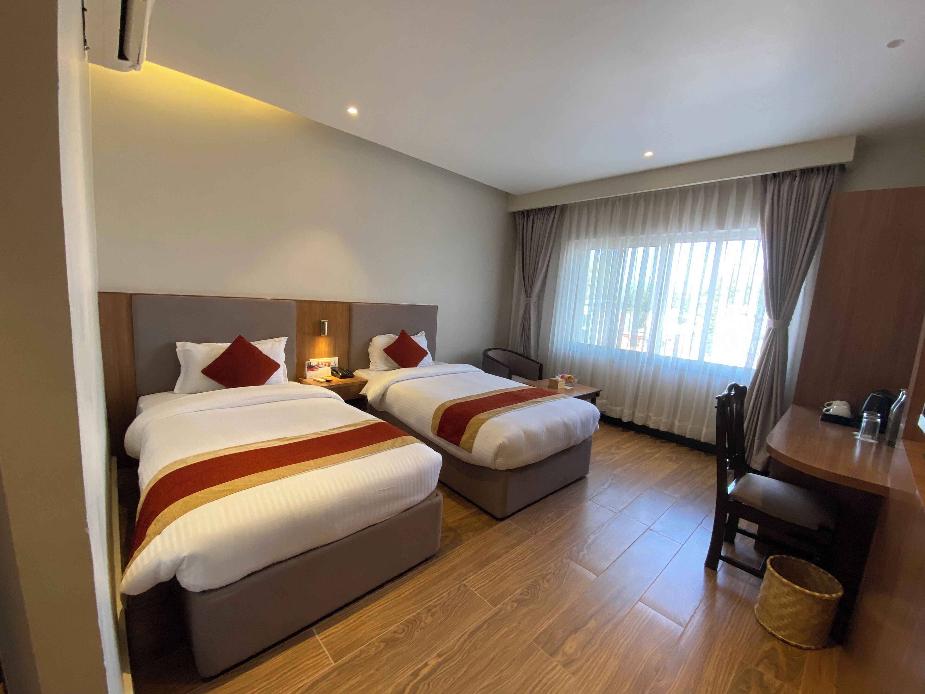 Quarto de hotel com duas camas, uma escrivaninha, uma cadeira e uma janela com cortinas.