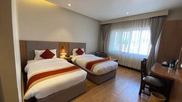 Quarto de hotel com duas camas, uma escrivaninha, uma cadeira e uma janela com cortinas.