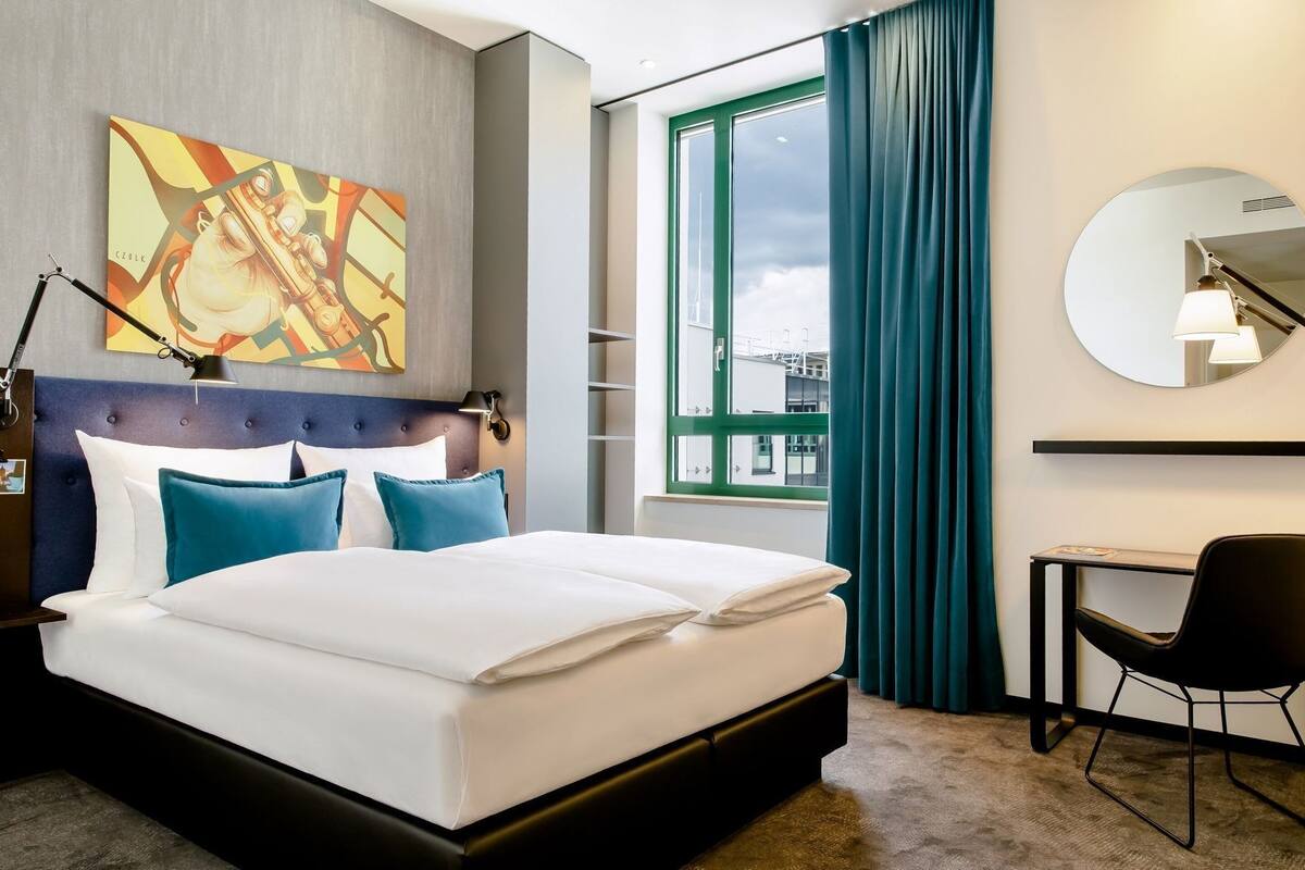 Quarto de hotel com cama, escrivaninha, cadeira, janela grande com cortinas, luminária na parede e um quadro.