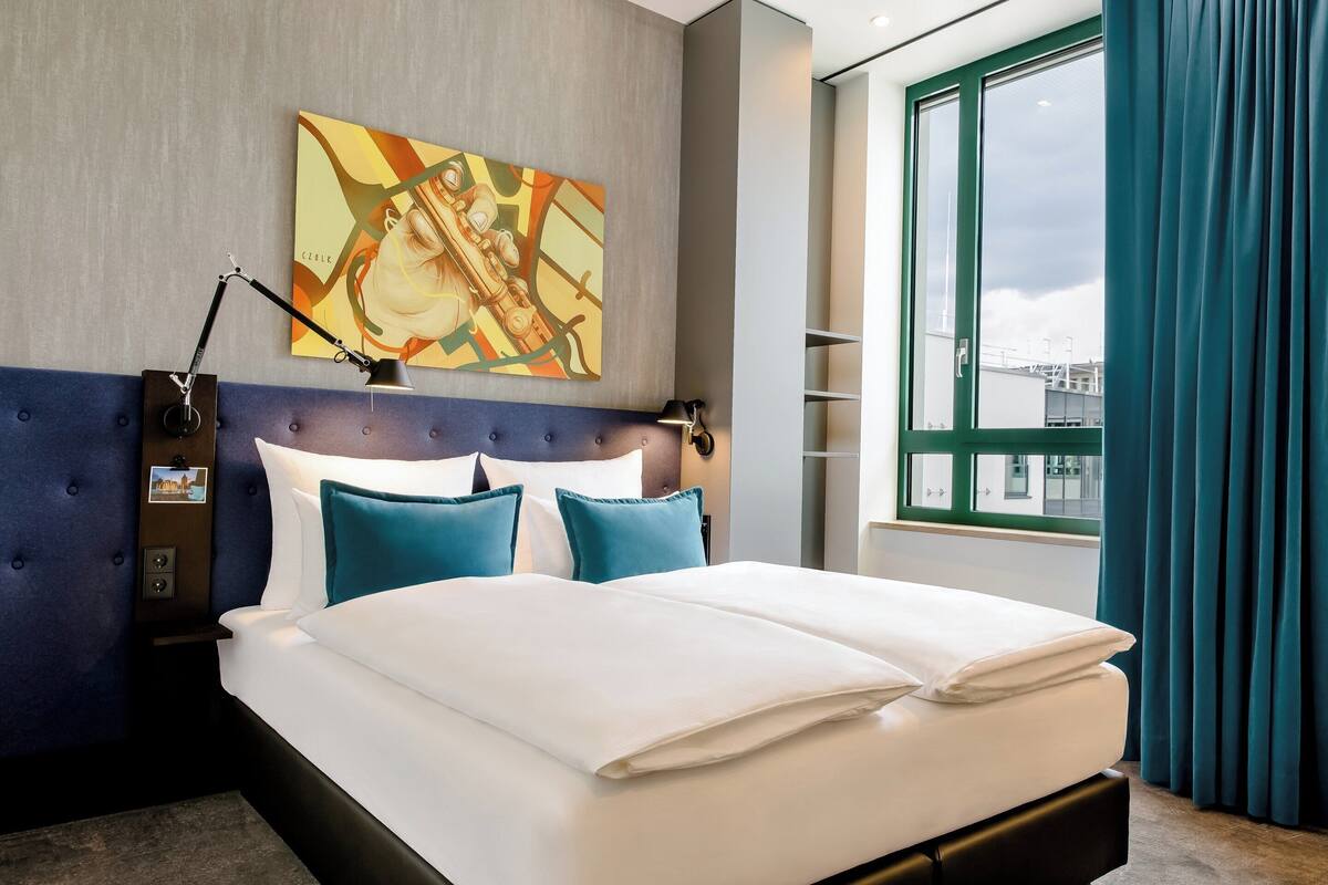 Quarto de hotel moderno com uma cama grande, cabeceira com prateleira embutida, um quadro na parede e vista para a cidade pela janela.