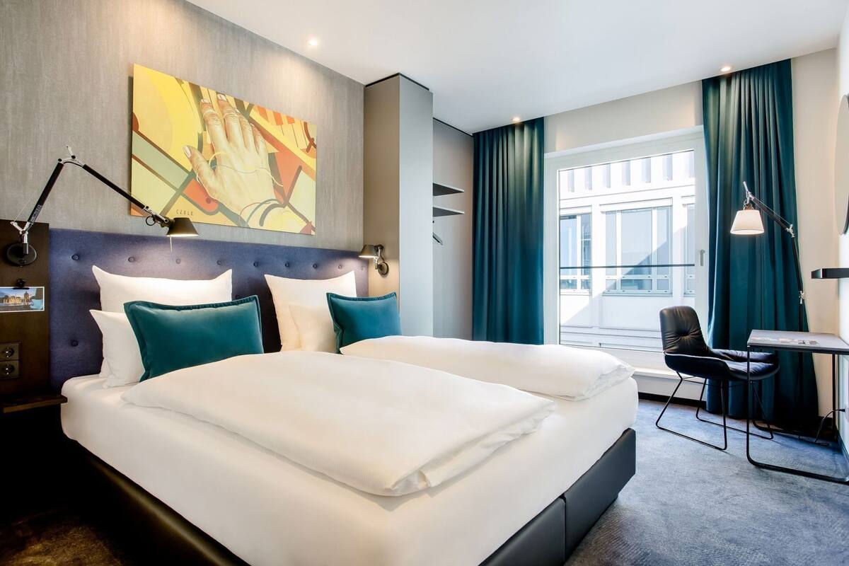 Quarto de hotel com uma cama grande, uma escrivaninha, uma cadeira, uma janela com cortinas e um quadro na parede.