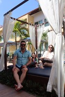 Um casal e uma criança sentados em um banco sob um toldo em ambiente externo, com palmeiras e um edifício ao fundo.