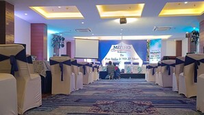 Uma sala de conferências com fileiras de cadeiras, palco com um banner e piso coberto de carpete.