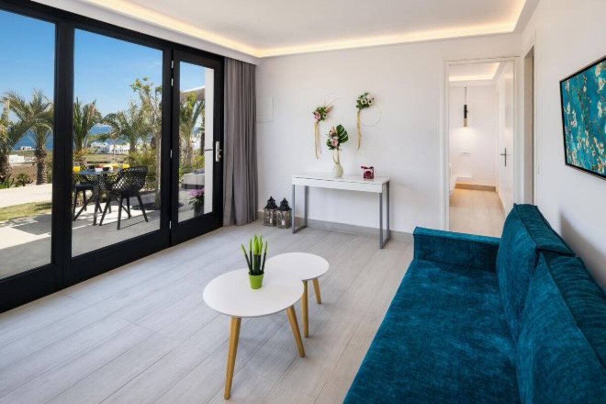 Uma sala de estar moderna com uma grande porta de vidro deslizante, um sofá azul, uma mesa redonda branca com duas cadeiras, um aparador branco com objetos decorativos e um quadro na parede.