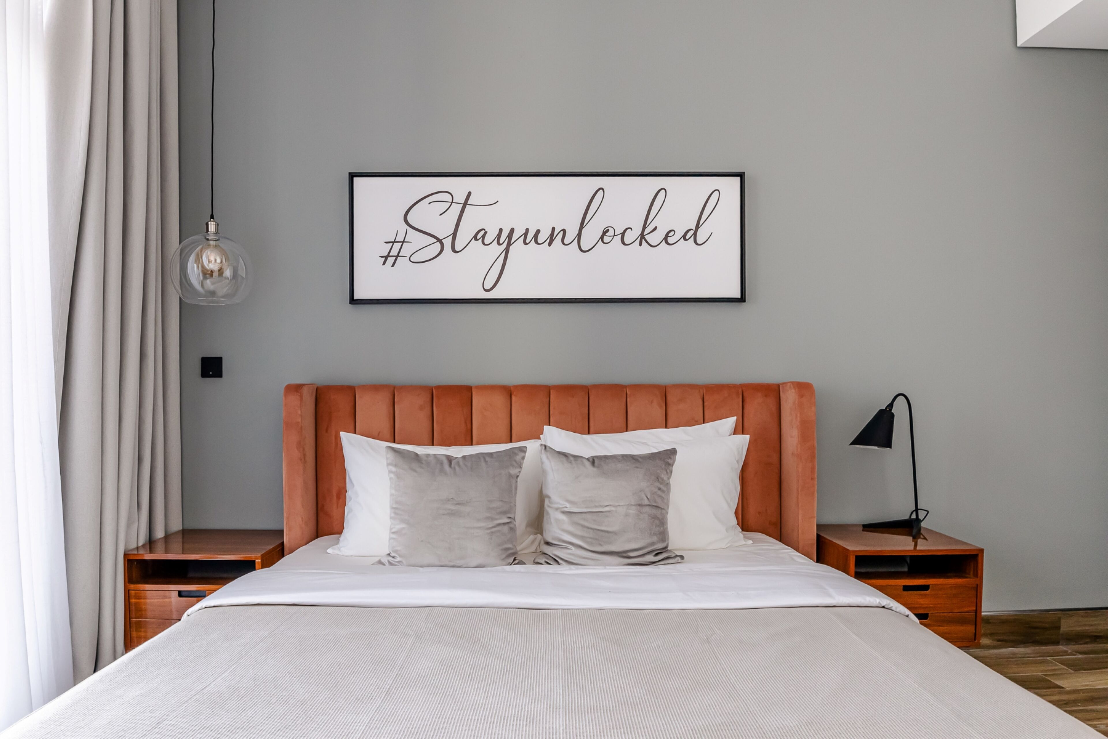 Uma cama bem arrumada com cabeceira, duas mesas de cabeceira, um aplique de parede e uma placa emoldurada com o texto #Stayunlocked.