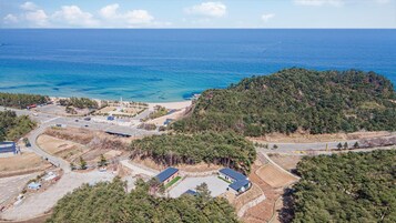 Vista aérea de uma área costeira com uma praia, uma pequena cidade e uma colina arborizada.