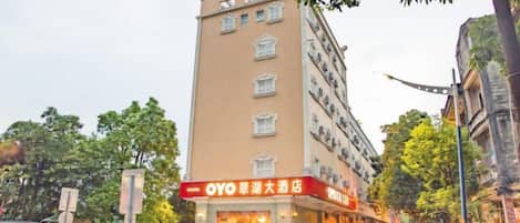 Um hotel com uma placa vermelha chamativa, vários andares e localização de esquina, com árvores e carros estacionados.