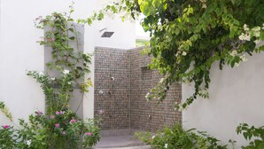 Pequena área de chuveiro externa, com parede de azulejos e degraus de acesso, cercada por vasos com plantas e flores.