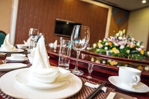 Uma mesa de jantar bem posta, com pratos, copos e uma xícara, em um ambiente com painéis de madeira e uma televisão ao fundo.