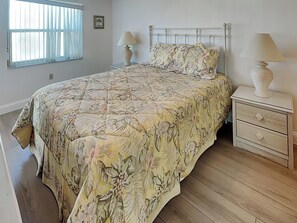 Quarto com colcha florida, cabeceira de metal branco, duas luminárias de cabeceira e mesa de cabeceira com gaveta.