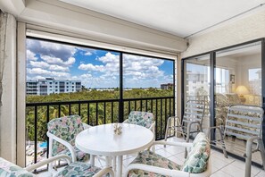Varanda com mesa branca e cadeiras florais, com vista para uma paisagem urbana com edifícios e céu claro.