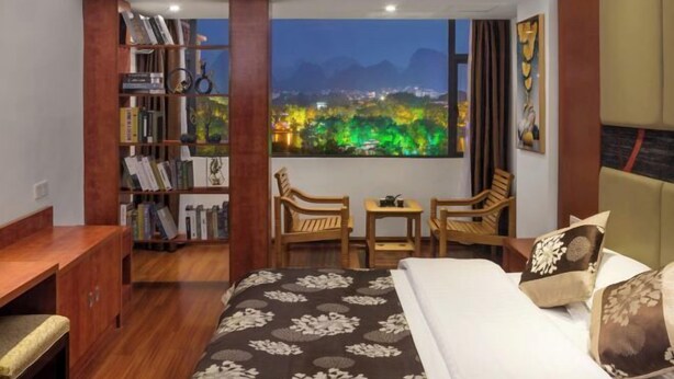 Quarto de hotel com uma cama grande, uma estante, uma escrivaninha, uma cadeira, uma mesa pequena e vista para a cidade à noite.