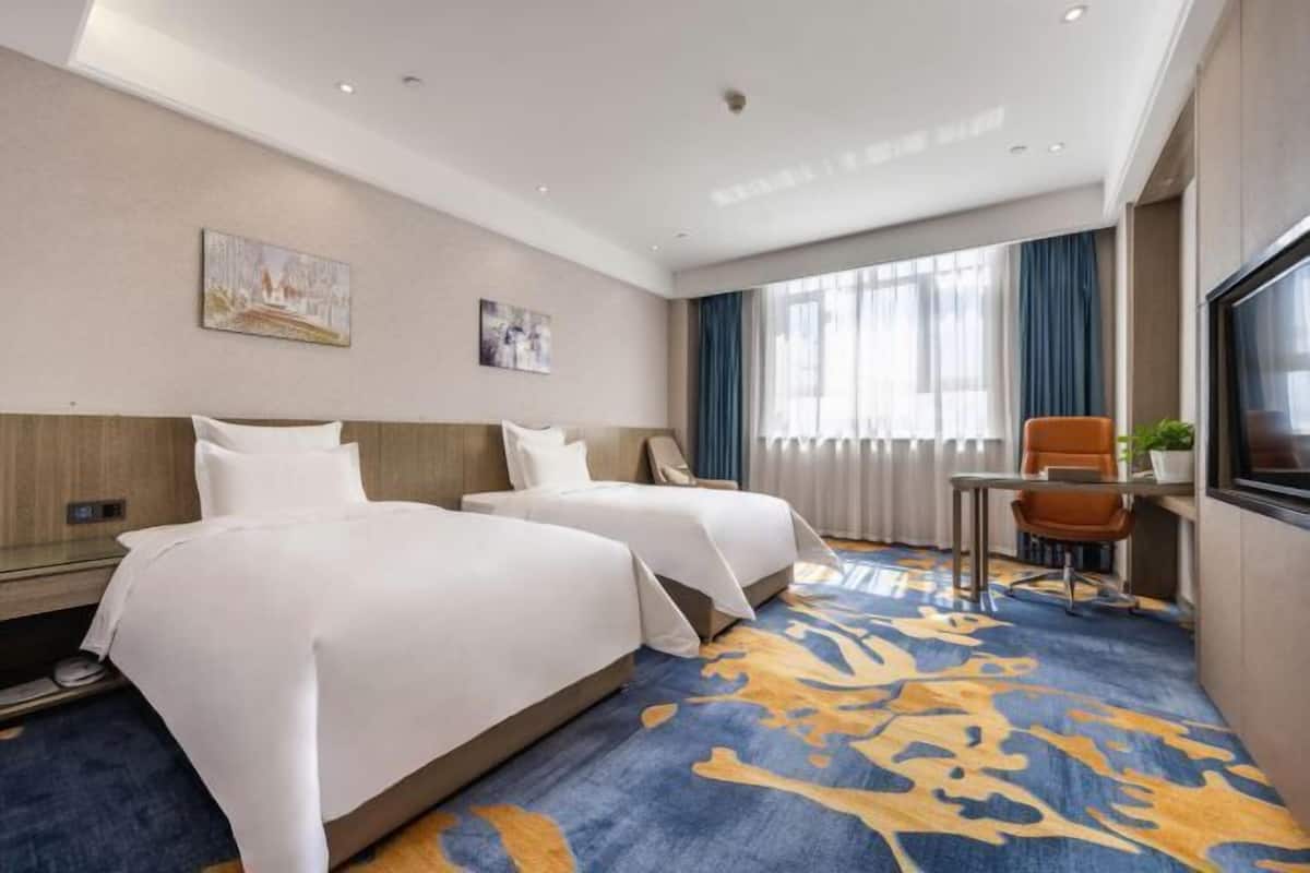 Quarto de hotel com duas camas, uma escrivaninha, uma cadeira e uma janela grande com cortinas.