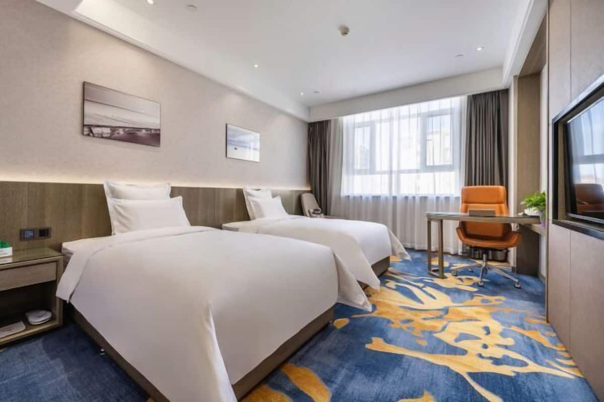 Quarto de hotel com duas camas, uma escrivaninha, uma cadeira e uma janela grande.