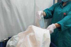 Uma pessoa vestida com uniforme médico segurando um pano branco, em pé num quarto com cortina.