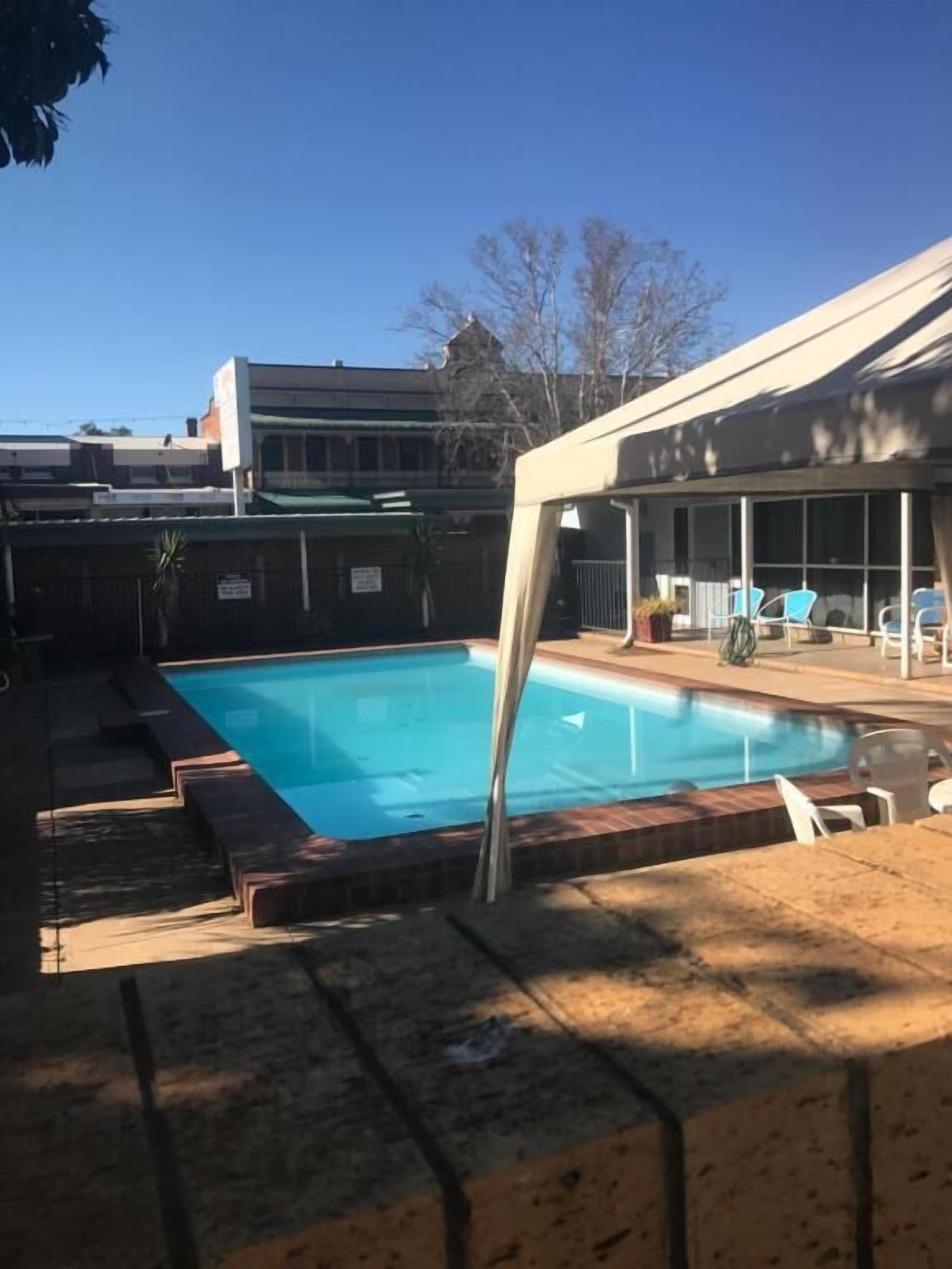 Piscina com área de estar coberta, quintal cercado e um edifício ao fundo.