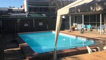 Piscina com área de estar coberta, quintal cercado e um edifício ao fundo.