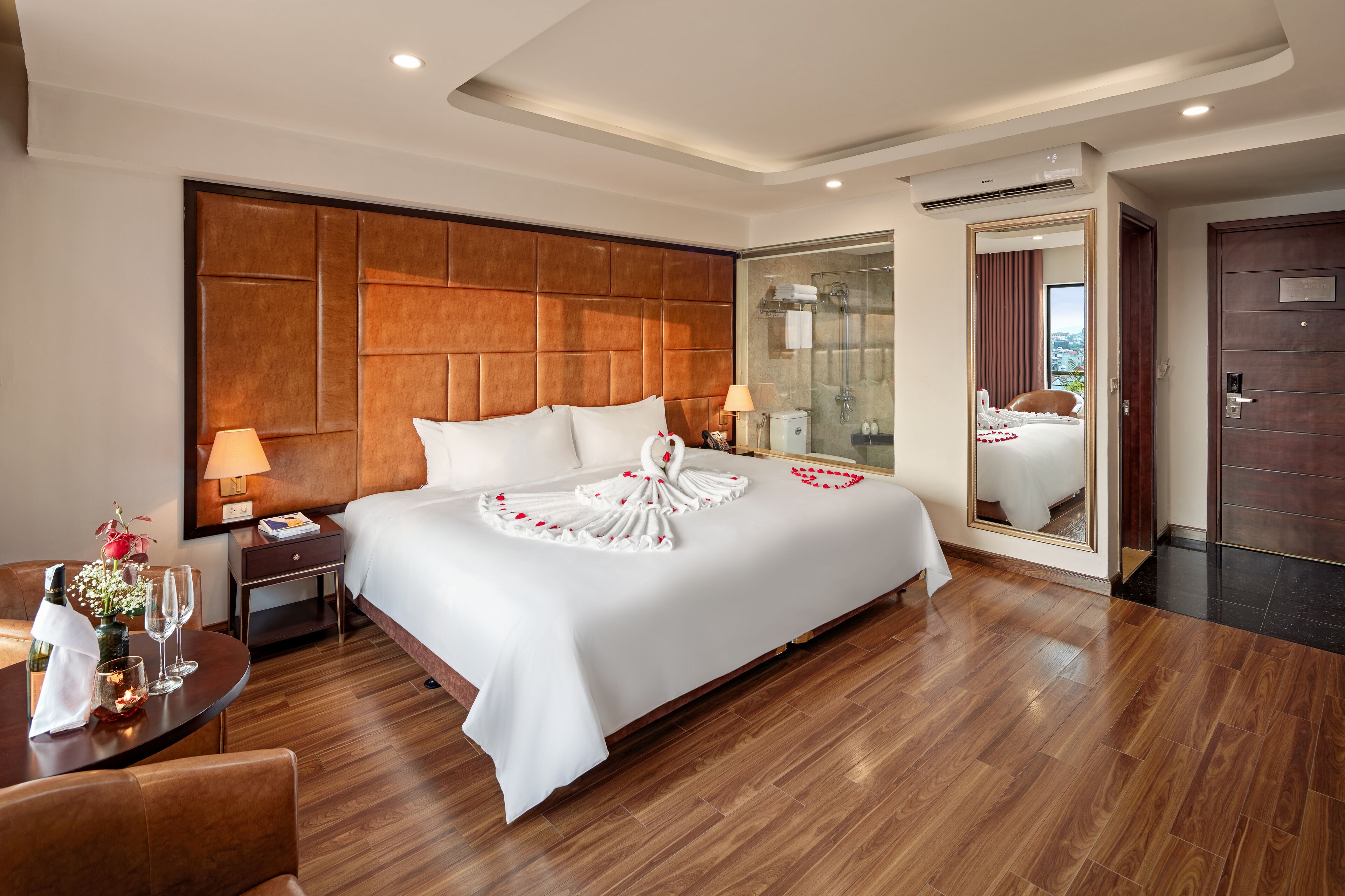 Quarto de hotel com uma cama grande, cabeceira de madeira, box de chuveiro de vidro e piso de madeira.